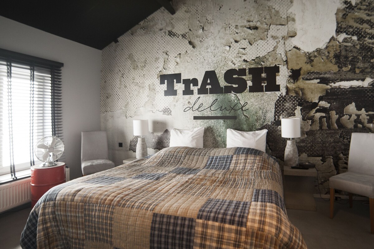 Quarto com uma cama, duas cadeiras, um ventilador e uma parede com a placa 'TRASH'.