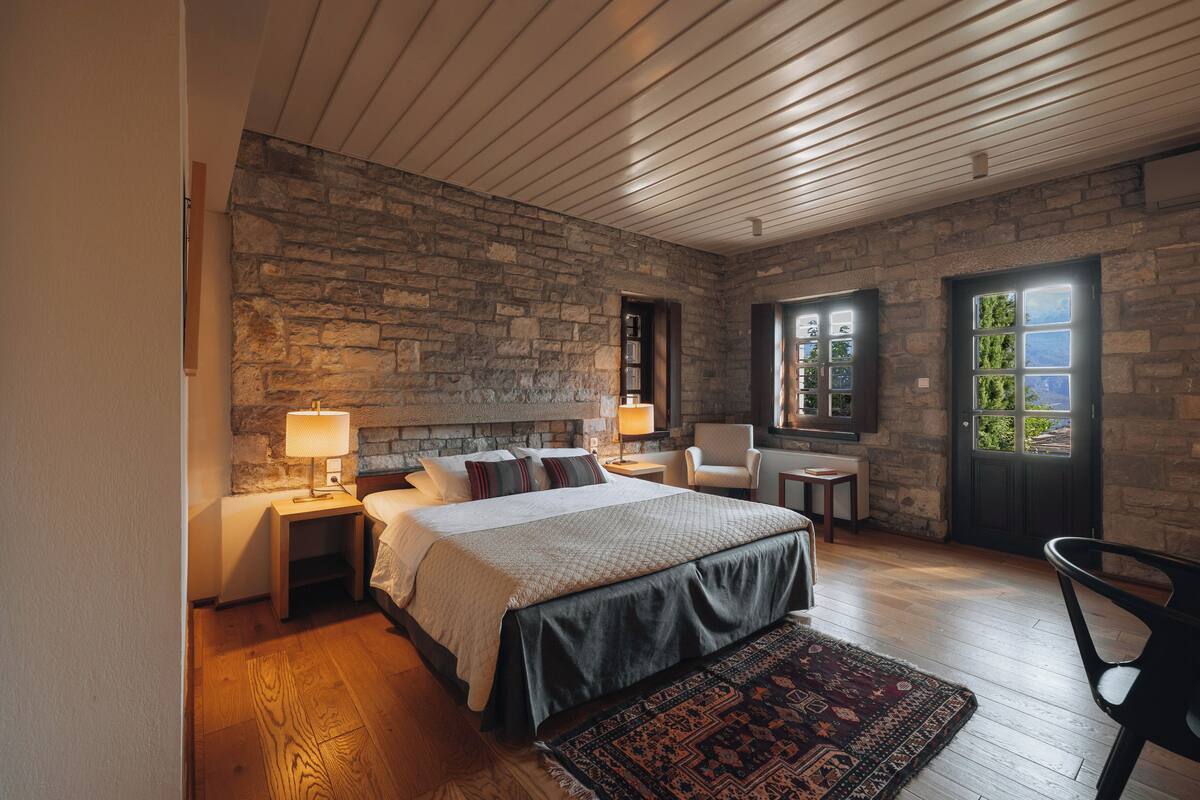 Quarto com parede de pedra, teto de madeira, cama com colcha escura, uma mesinha de cabeceira com abajur e uma cadeira.
