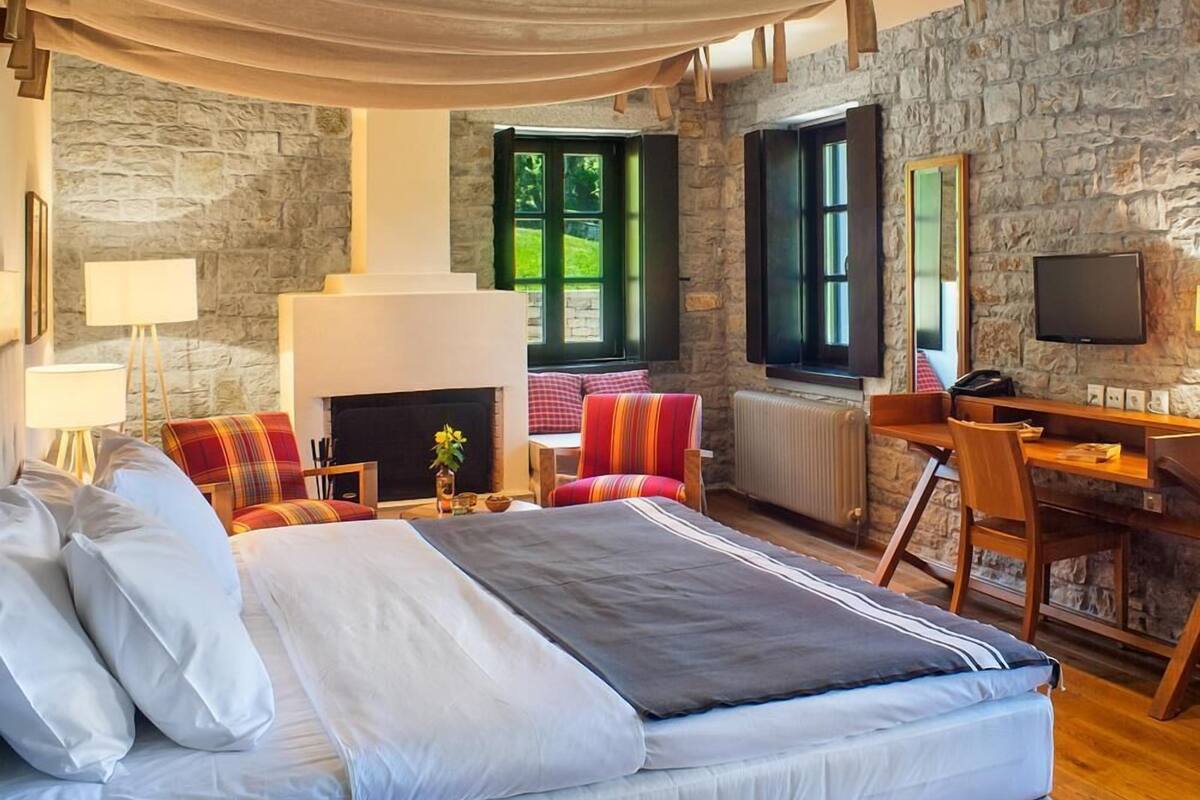 Um quarto com cama, escrivaninha, poltrona, lareira e parede de pedra.