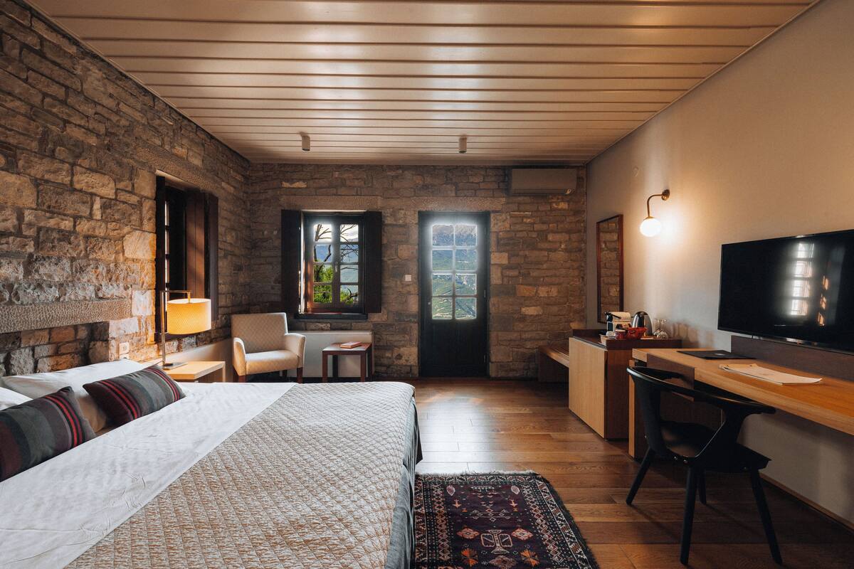 Quarto de hotel com uma cama grande, escrivaninha, cadeira, televisão e vista para o exterior pela janela.