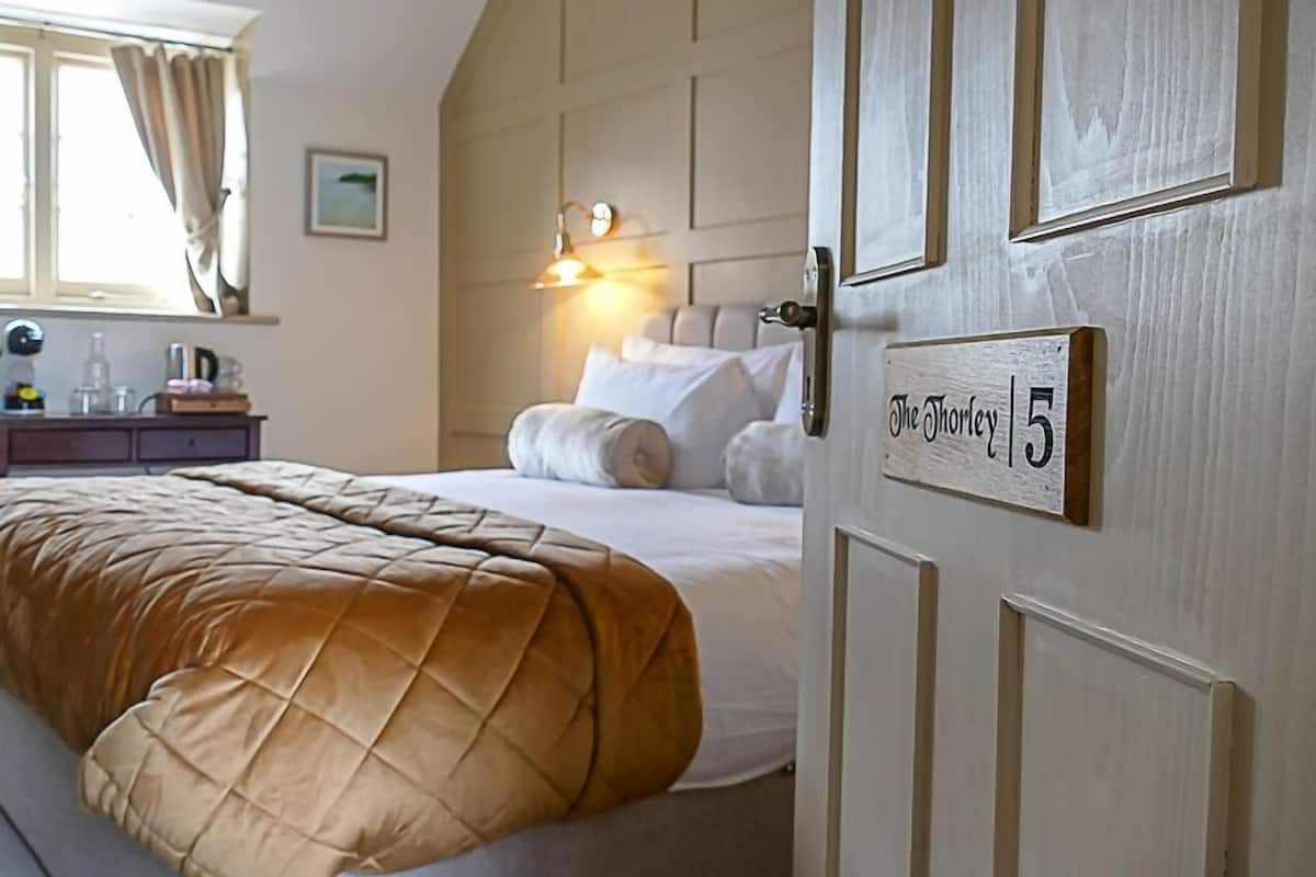 Quarto com cama, mesa de cabeceira, janela com cortinas, placa na parede e porta.