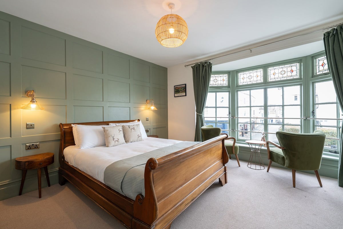 Quarto com cama de madeira, duas mesinhas de cabeceira, poltrona verde, mesa pequena e uma janela grande com cortinas.