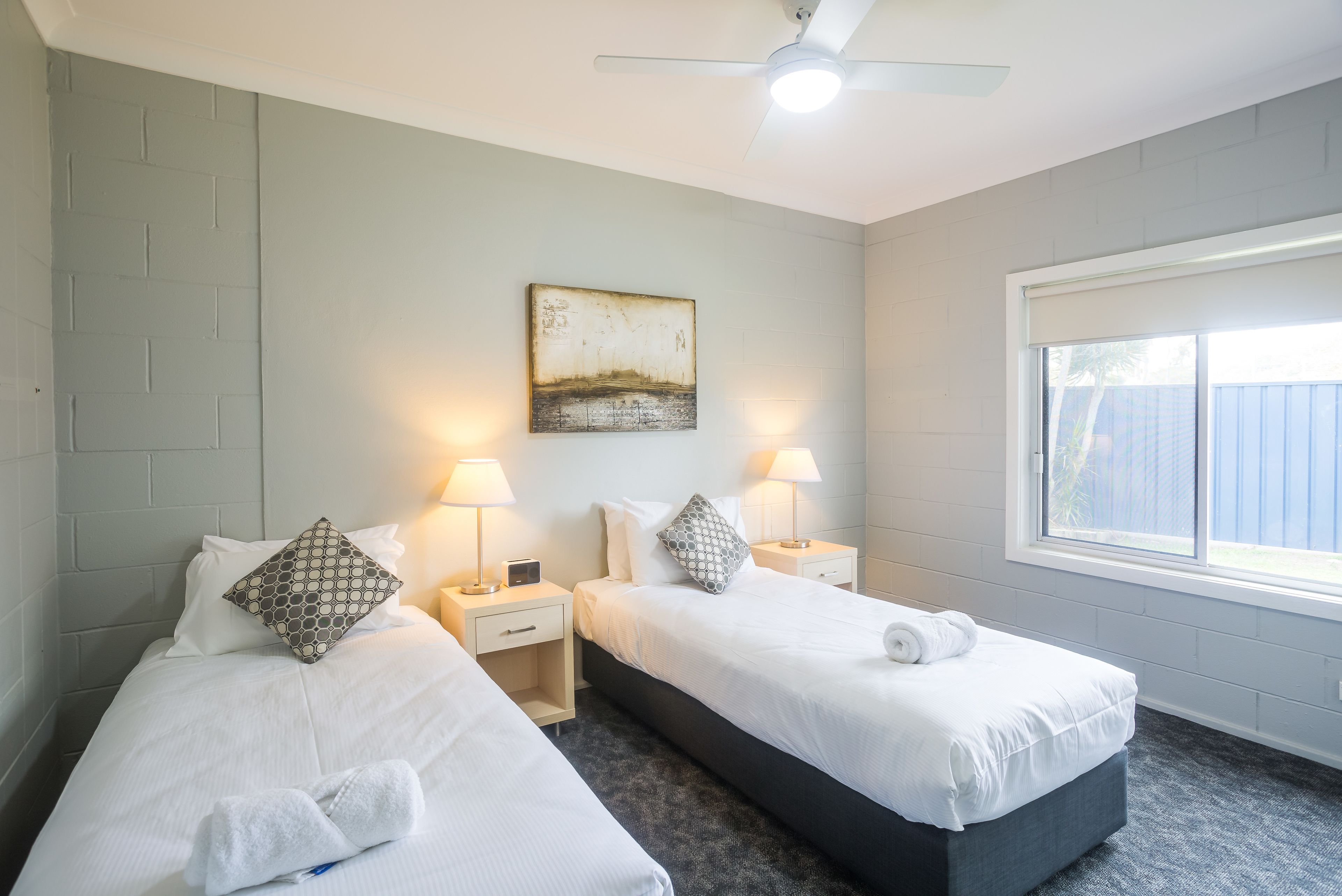 Quarto de hotel com duas camas, um ventilador de teto, uma janela com vista e um quadro na parede.
