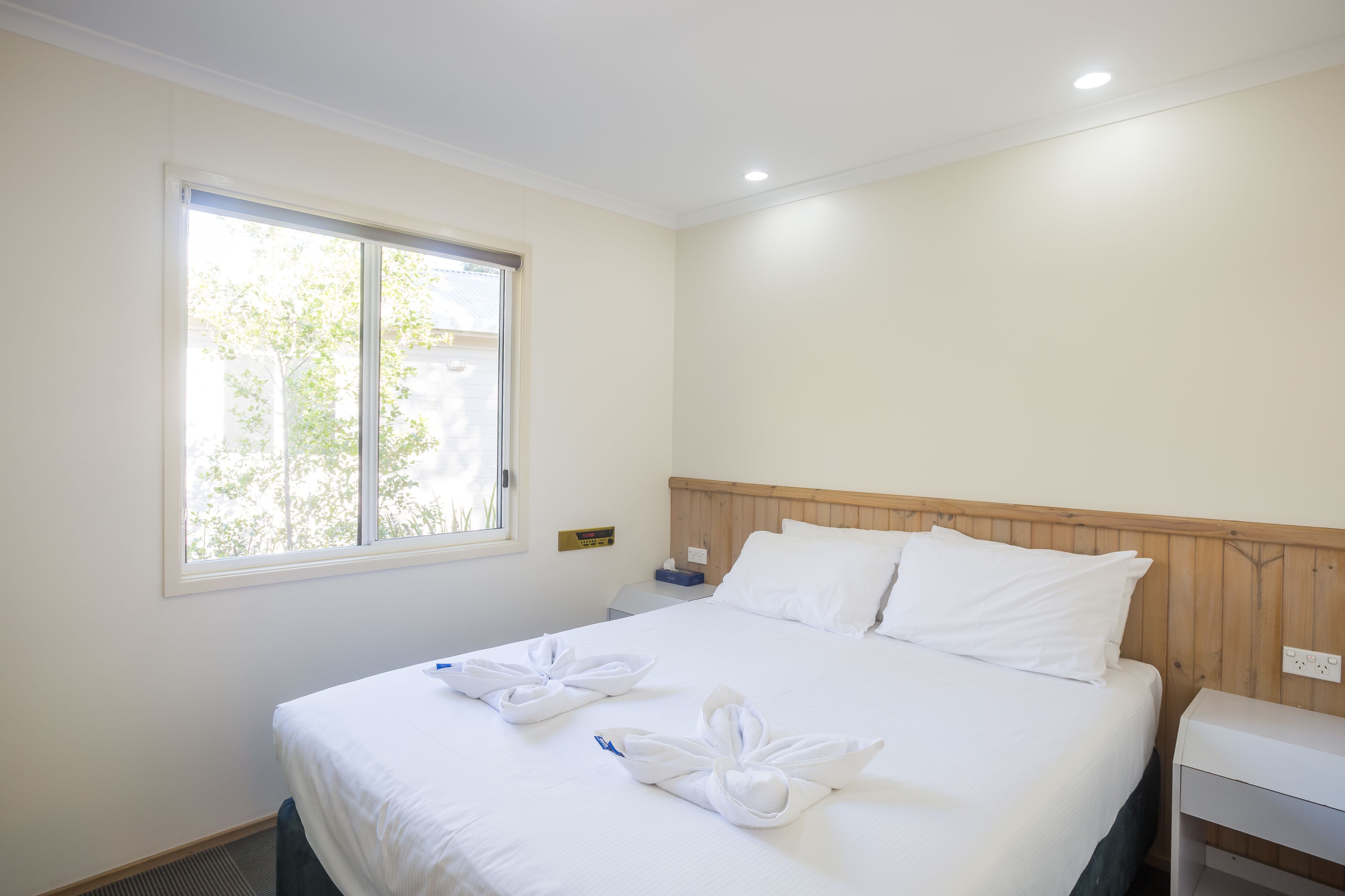 Quarto de hotel com uma cama grande, cabeceira de madeira, mesa de cabeceira e janela com vista para árvores.