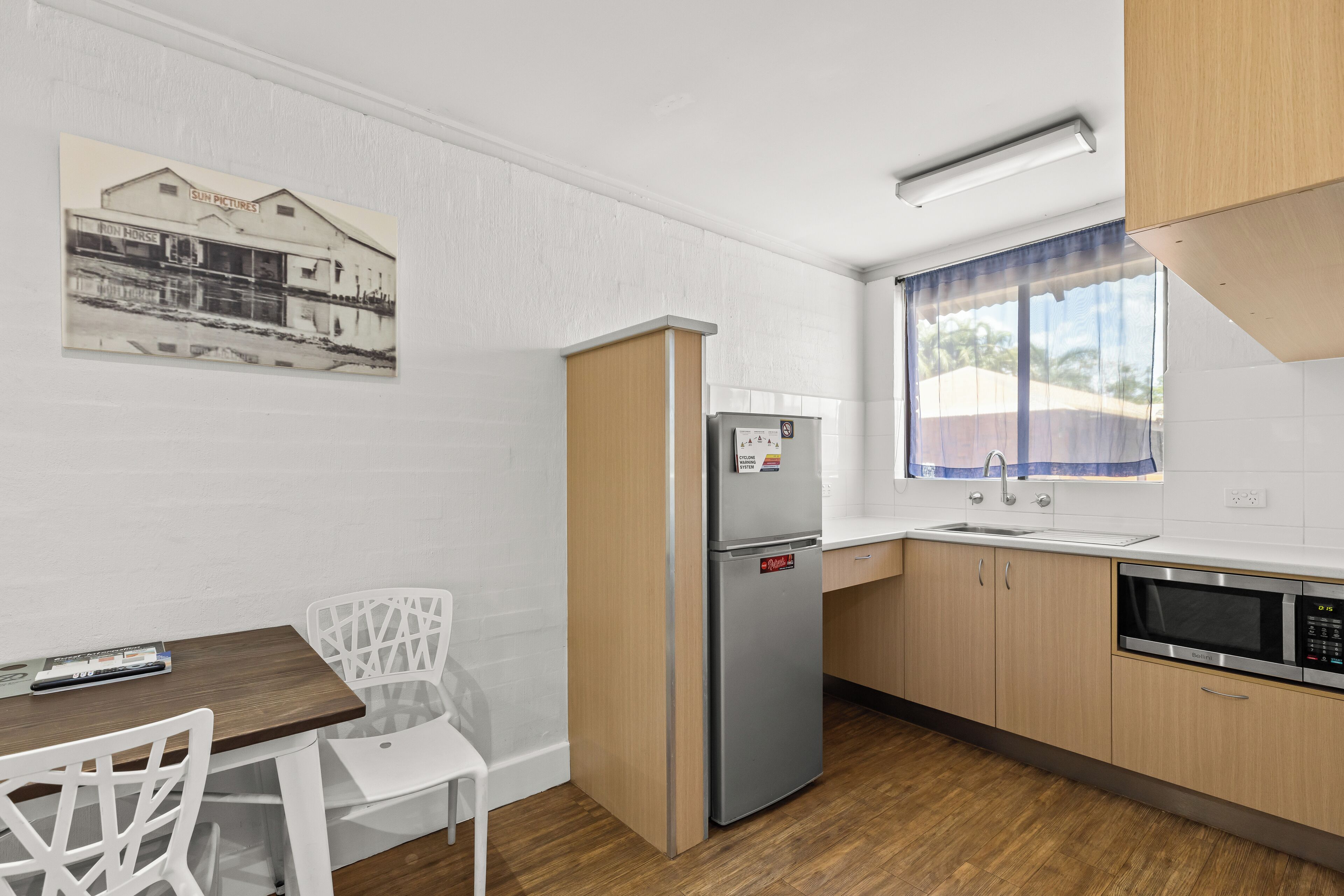 Uma cozinha compacta com geladeira, micro-ondas e pia. Há uma área de refeições com mesa e cadeiras. Uma janela com cortina é visível, e há um quadro fixado na parede.