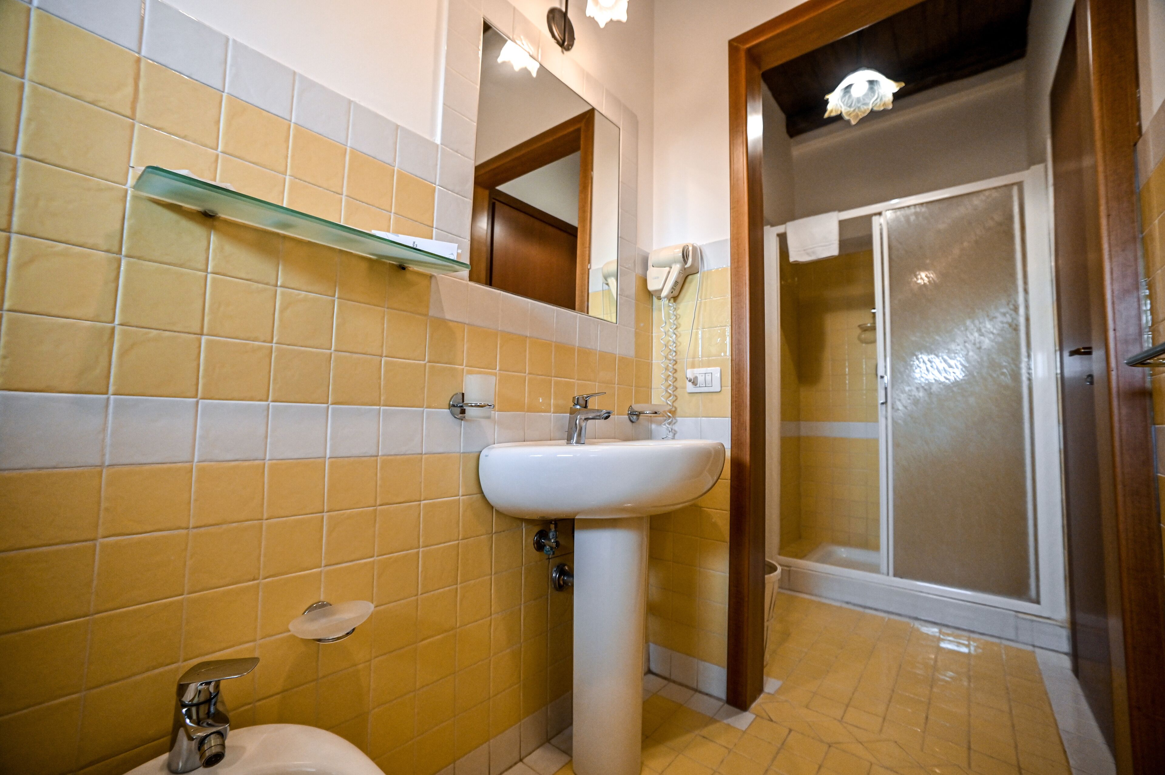Uma casa de banho com azulejos amarelos e brancos, uma pia branca, uma estante de vidro, um espelho e uma área de duche.