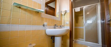 Uma casa de banho com azulejos amarelos e brancos, uma pia branca, uma estante de vidro, um espelho e uma área de duche.