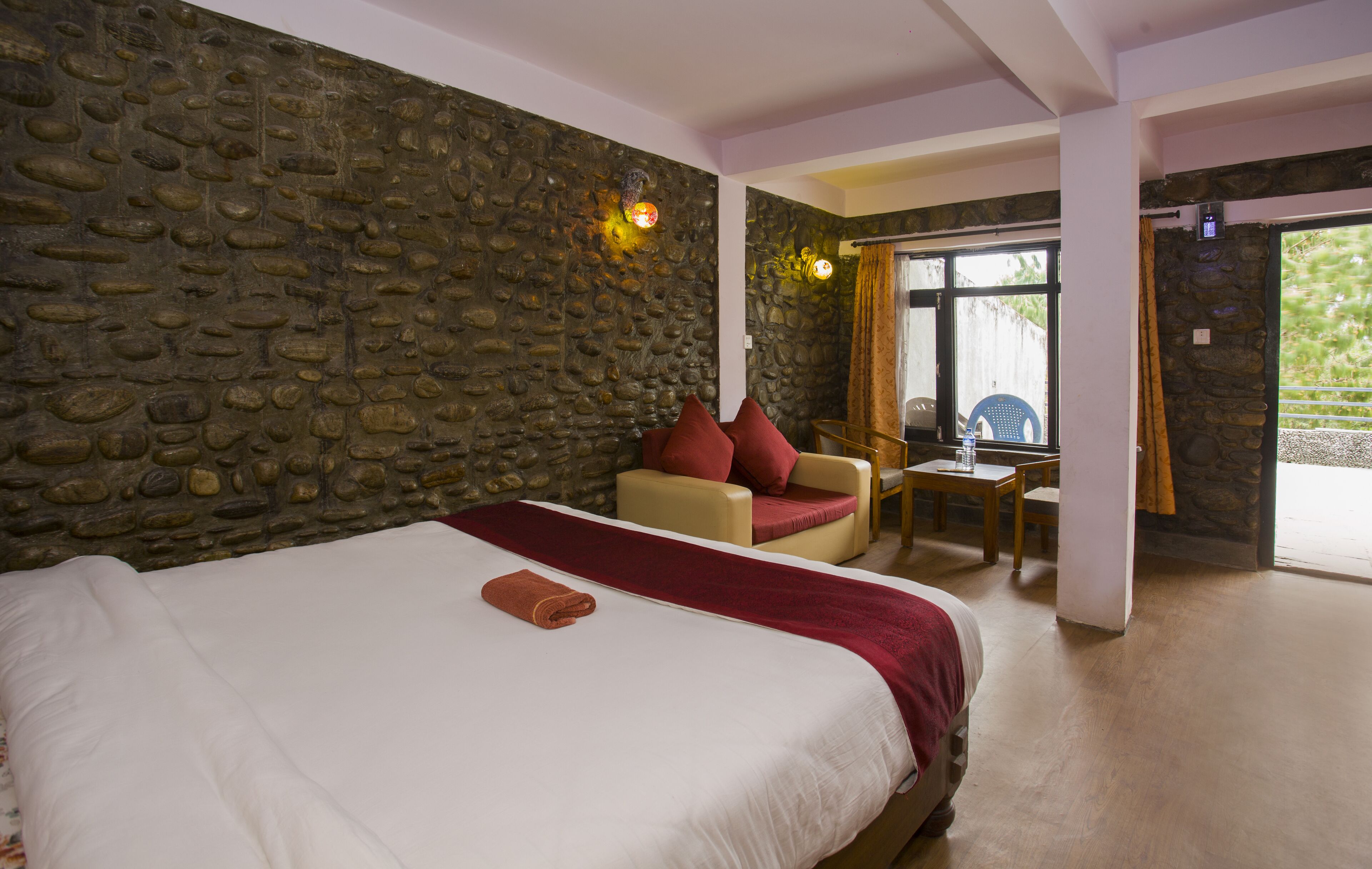 Quarto de hotel com parede de pedra, cama com manta vermelha, uma cadeira, uma mesa pequena e vista para o exterior.