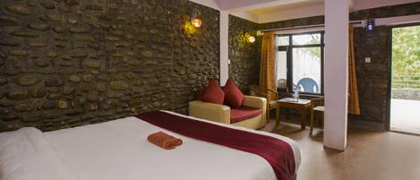 Quarto de hotel com parede de pedra, cama com manta vermelha, uma cadeira, uma mesa pequena e vista para o exterior.