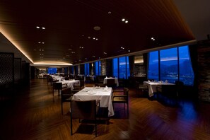 Restaurante com mesas postas para refeições, amplas janelas com vista para a cidade e teto de madeira.