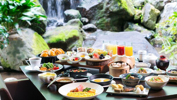 Buffet de café da manhã com variedade de pratos, incluindo ovos, carnes e frutas, com uma cascata e vegetação ao fundo.