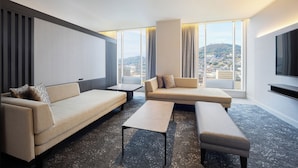 Quarto de hotel moderno com sofá, dois pufes, mesa de centro e televisão. O quarto possui amplas janelas com vista para a cidade.