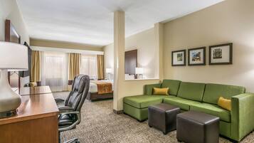 Quarto de hotel com um sofá verde, uma mesa com abajur, uma cadeira e uma cama com cabeceira branca. Há três quadros na parede.