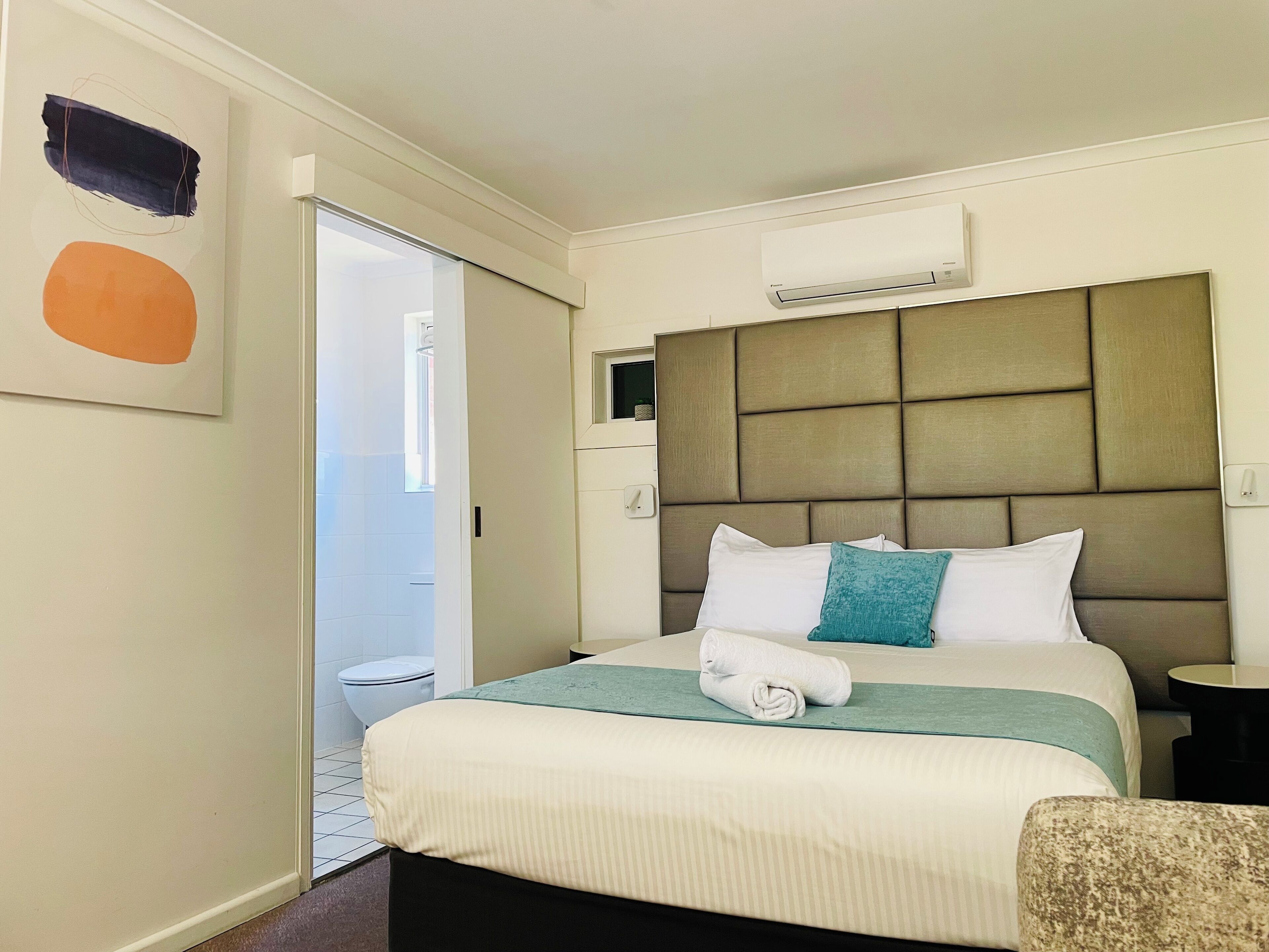 Quarto de hotel com cama, mesa de cabeceira, ar-condicionado na parede e porta de acesso a um banheiro.