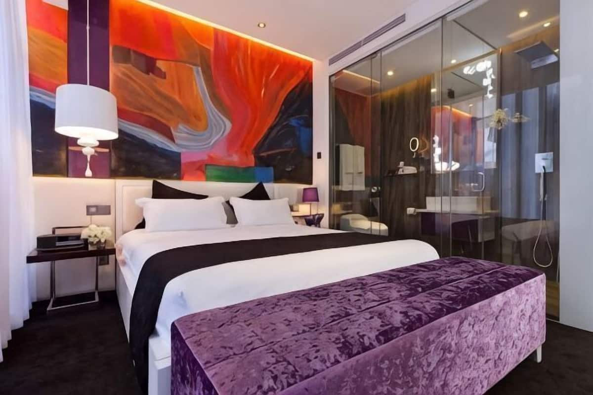 Quarto de hotel moderno com uma cama grande, uma mesa de cabeceira, um criado-mudo, chuveiro box envidraçado e um mural abstrato vibrante na parede.