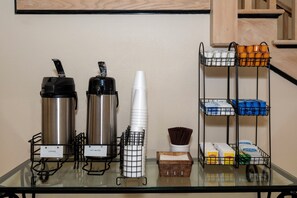 Estação de autoatendimento com dispensadores de café e água quente, copos de papel e diversos artigos de higiene pessoal nas prateleiras.