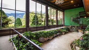 Amplo espaço interno com grandes janelas que oferecem vista para as montanhas, uma escada e diversas plantas.