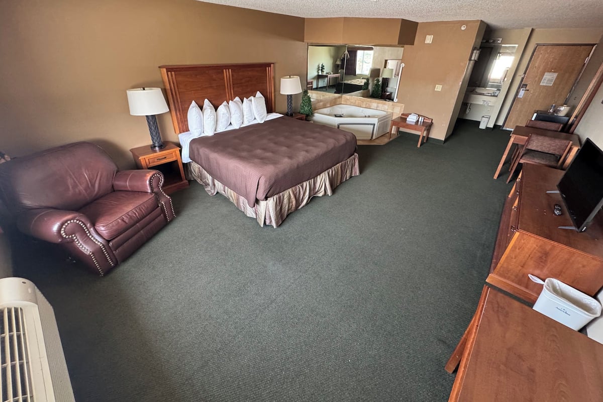 Quarto de hotel com uma cama grande, uma poltrona de couro, uma escrivaninha com um computador, uma pequena mesa com abajur e um banheiro visível através de uma porta.