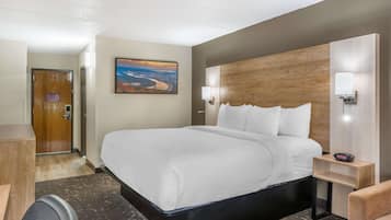 Quarto de hotel com uma cama grande, uma escrivaninha, uma cadeira, uma mesinha de cabeceira e um quadro na parede.
