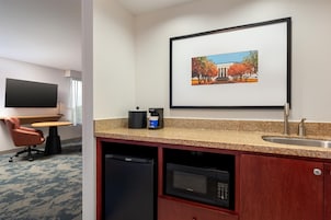 Quarto de hotel com cozinha compacta, televisão de tela plana, quadro pendurado na parede e escrivaninha com cadeira.