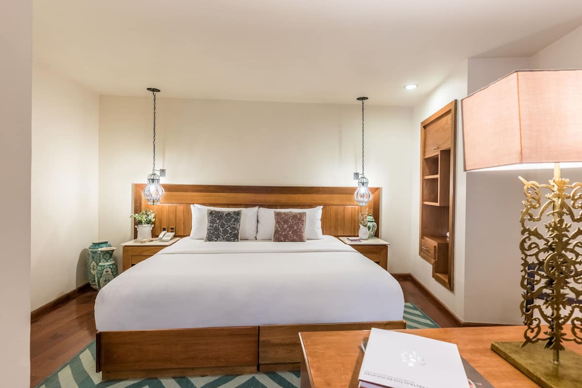Quarto de hotel com uma cama grande, cabeceira de madeira, duas mesas de cabeceira com luminárias, um criado-mudo, um vaso decorativo e um guarda-roupa de madeira.