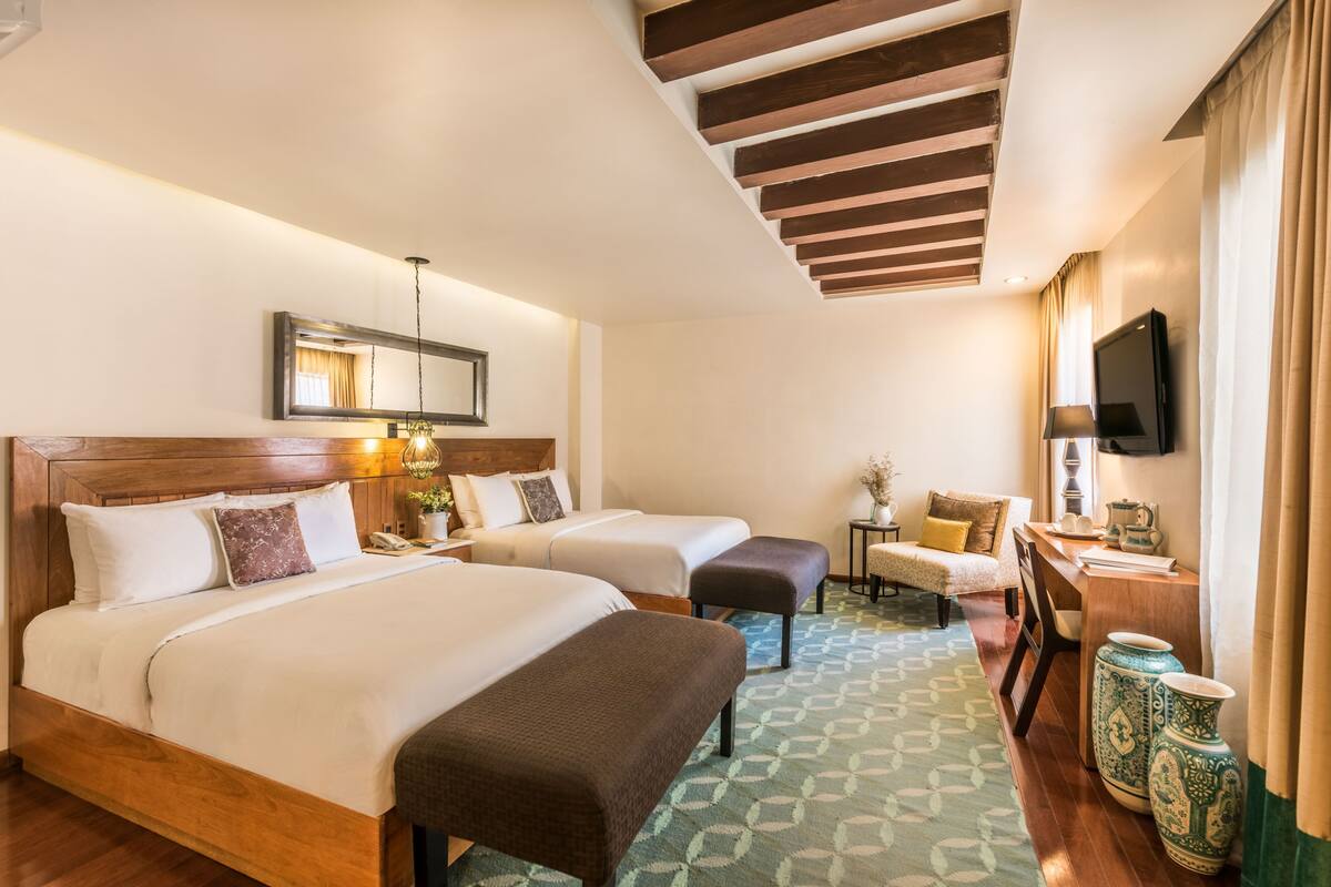 Quarto de hotel com duas camas, uma escrivaninha, uma cadeira, uma televisão e um tapete estampado.