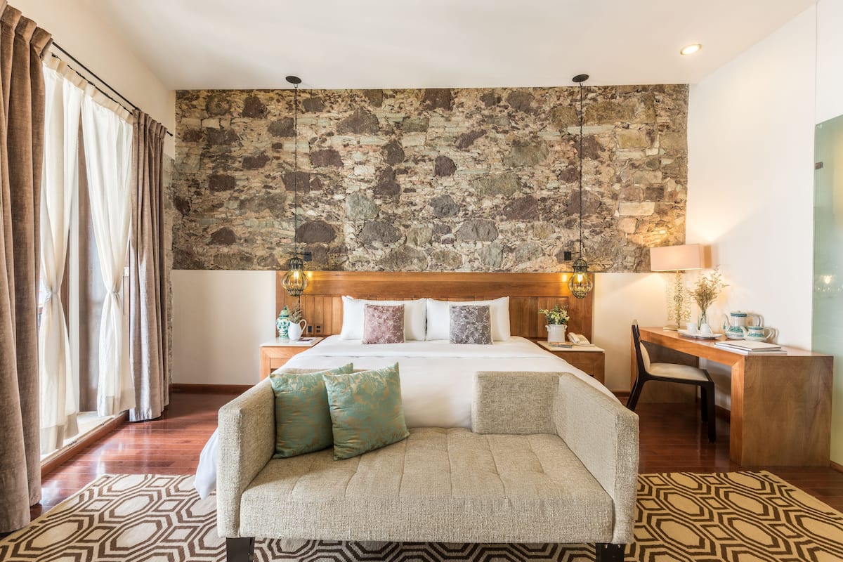 Quarto de hotel com parede de pedra, cama com várias almofadas, sofá, escrivaninha e abajur.