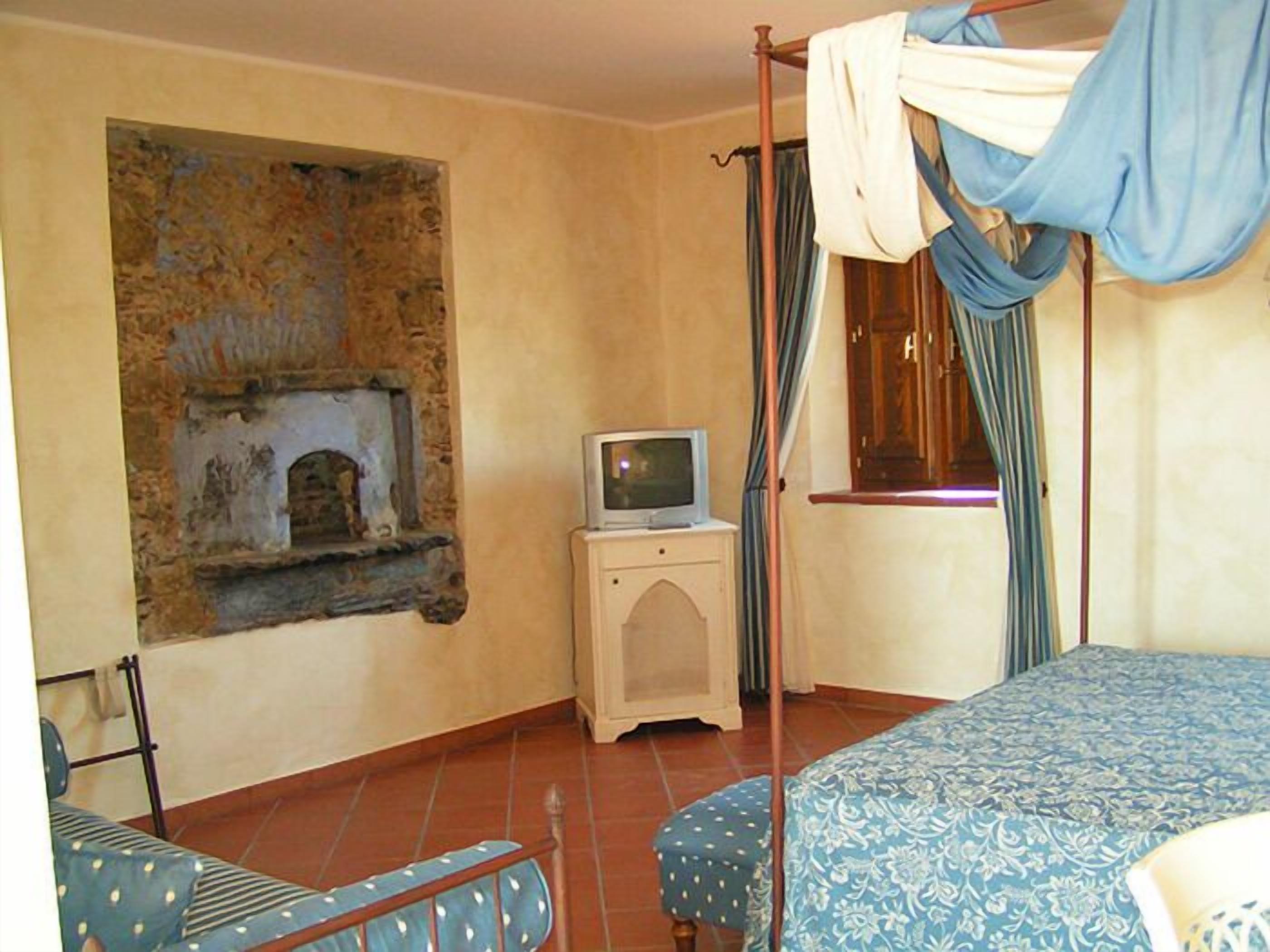 Quarto com lareira de pedra, televisão sobre uma mesinha, cama com dossel e janela com cortinas.