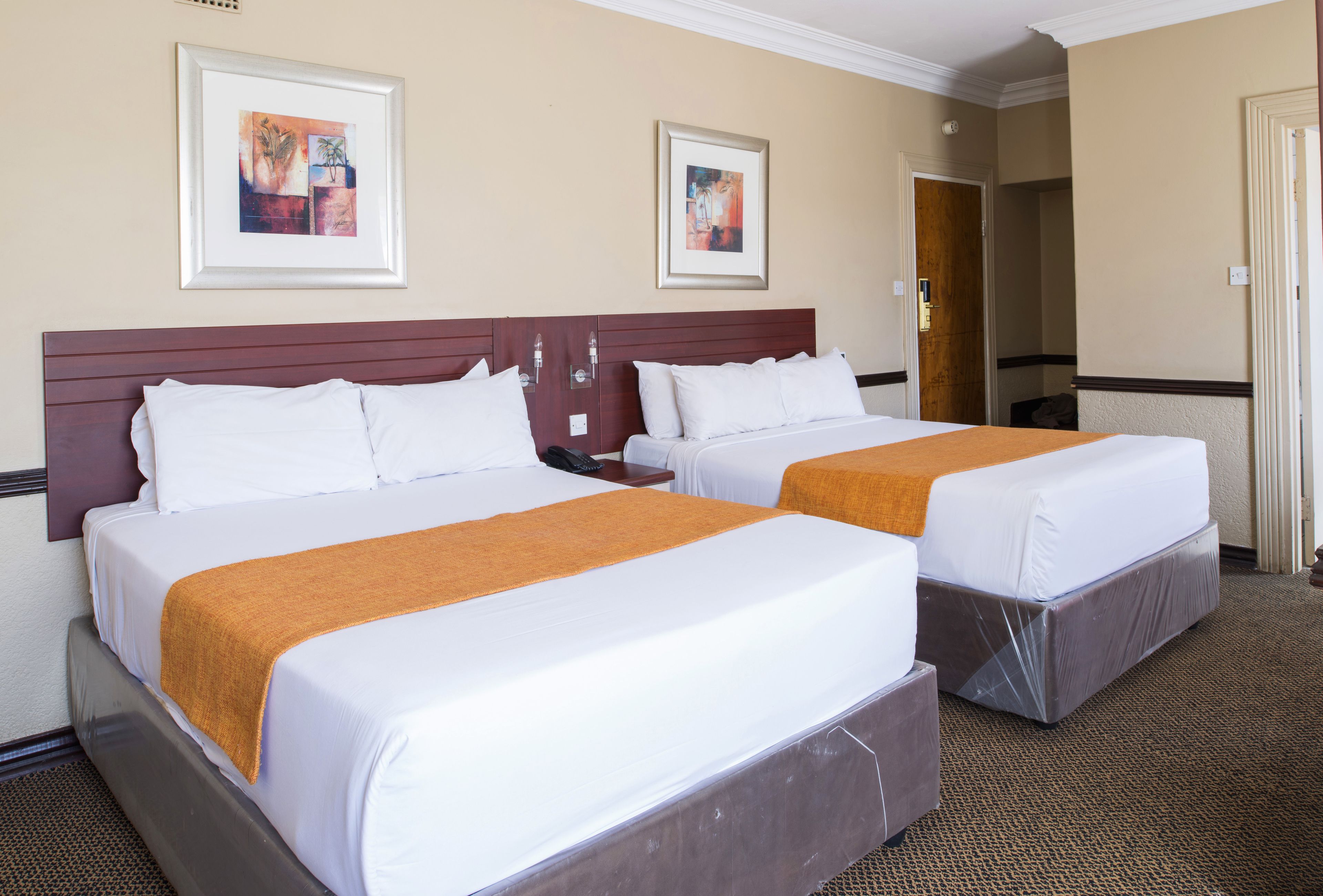 Quarto de hotel com duas camas, cada uma com roupa de cama branca e cabeceira marrom. Há quadros nas paredes e uma porta que dá para outro quarto.