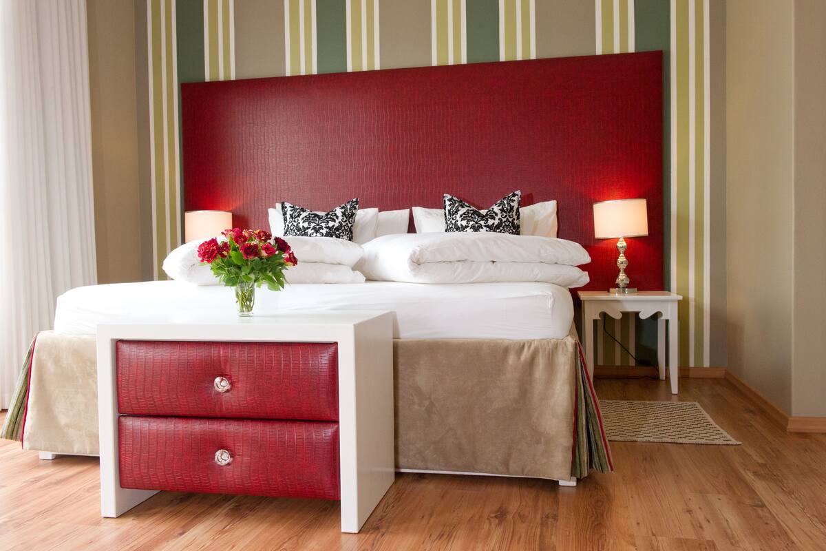 Quarto de hotel com uma cama grande, roupa de cama branca, cabeceira vermelha, duas mesas de cabeceira com abajures, uma mesinha de apoio vermelha e um pequeno vaso com flores.