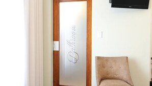 A bathroom with a frosted glass door labeled "Bathroom," a beige upholstered chair, and a mounted television on the wall.