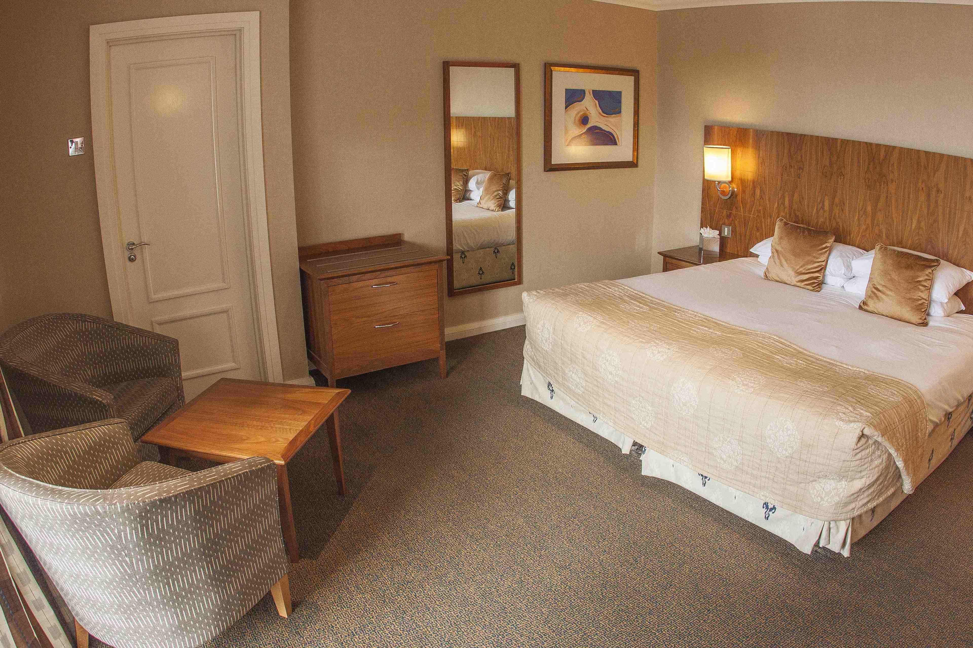 Quarto de hotel com cama, mesa de cabeceira de madeira, penteadeira, cadeira e pequena mesa.