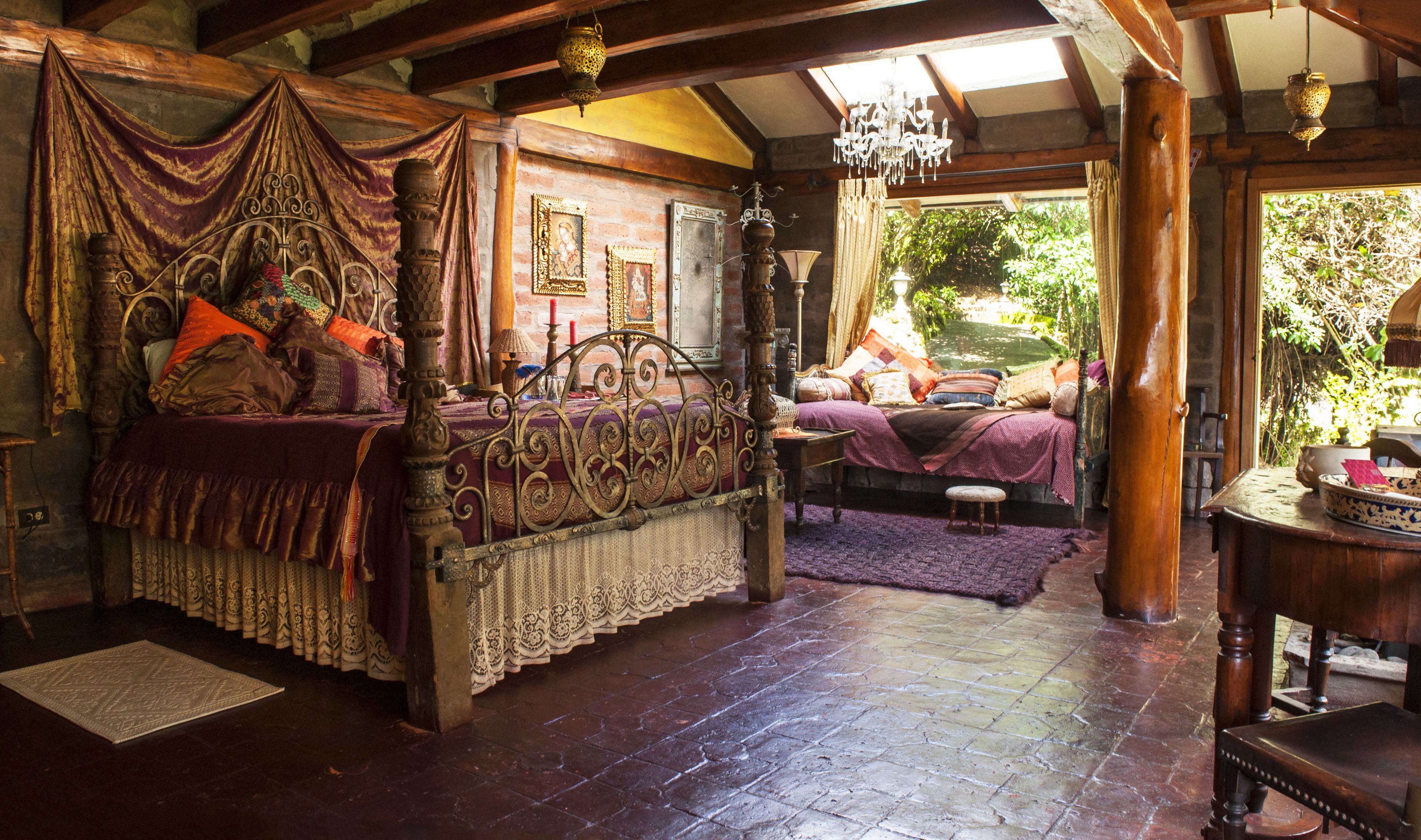 Uma cama de madeira tradicional com dossel e ornamentos elaborados, um candelabro e vista para um jardim exuberante através de uma porta aberta.