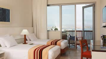 Quarto de hotel com duas camas, uma escrivaninha, uma cadeira e varanda com vista para o mar e barcos.