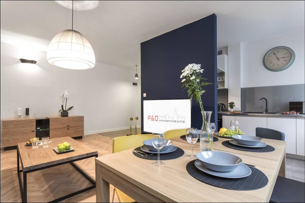 Uma zona de refeições moderna com uma mesa de madeira posta para quatro pessoas, um vaso com flores e um aviso a indicar Apartamentos P&O.