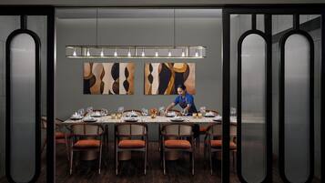 Uma sala de jantar com uma mesa comprida posta para seis pessoas, rodeada por cadeiras de vime com almofadas laranja. Uma pessoa está a pôr a mesa.