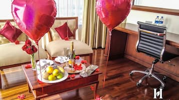 Quarto de hotel com uma cama, uma mesa com uma garrafa de champanhe, um prato com frutas e balões em formato de coração.