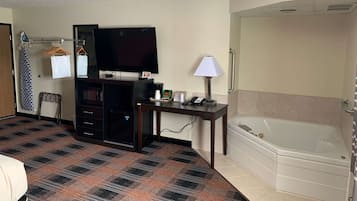 Quarto de hotel com carpete xadrez, uma mesa com luminária, televisão, minibar, cama e banheira.