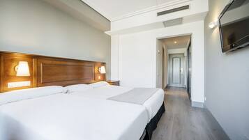 Quarto de hotel com cama de casal, luminárias de cabeceira, televisão de tela plana na parede e corredor que dá para outro quarto.