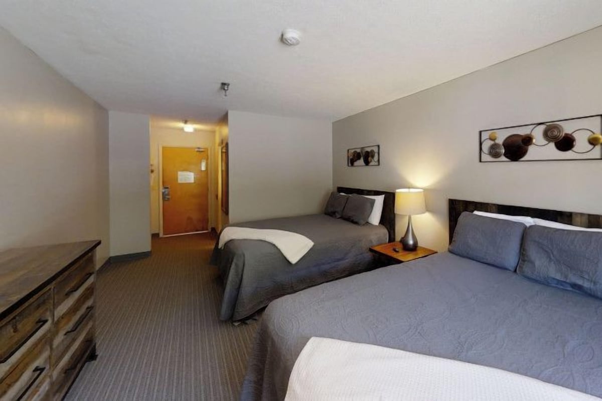 Quarto de hotel com duas camas, uma cômoda, um abajur e uma porta que dá para outro aposento.