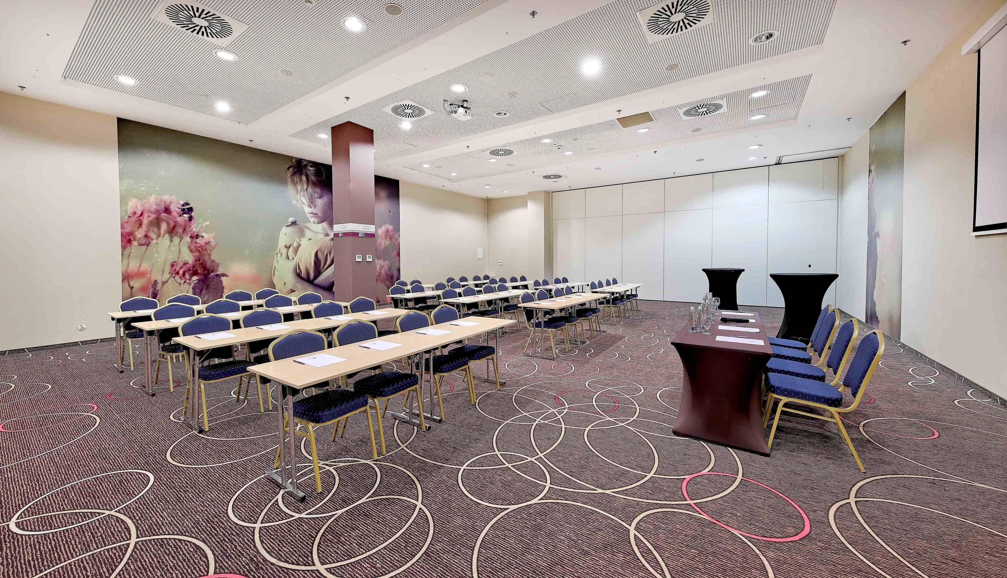 Sala de conferência com mesas e cadeiras dispostas em fileiras, um mural grande na parede e um tapete estampado.