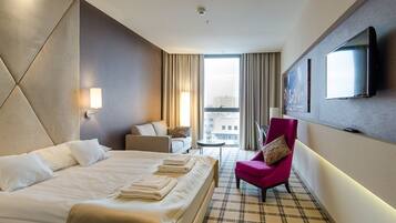 Quarto de hotel com uma cama grande, um sofá pequeno, uma cadeira, uma televisão e uma janela com cortinas.
