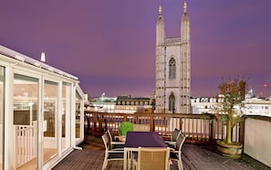 Terraço no telhado com mesa e cadeiras, com vista para uma torre de igreja histórica.