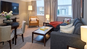 Quarto de hotel moderno com poltrona de couro, sofá cinza, mesa de centro com um livro, televisão e janela com cortinas.