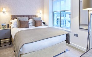Um quarto bem arrumado com uma cama grande, duas mesas de cabeceira, um criado-mudo, uma janela com vista e uma luminária fixada na parede.
