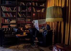 Um recanto aconchegante para leitura, com móveis de couro, uma estante repleta de livros, uma luminária e um jornal.