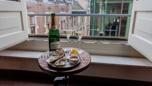 Uma mesa com uma garrafa de vinho, dois copos, ostras, limão e uma tigela de molho, junto a uma janela com vista para a cidade.