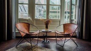 Quarto com duas poltronas de couro, uma mesa pequena com um vaso e vista para edifícios por amplas janelas.
