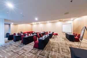 Sala de conferências com mesas, cadeiras, tela de projeção e piso coberto de carpete.