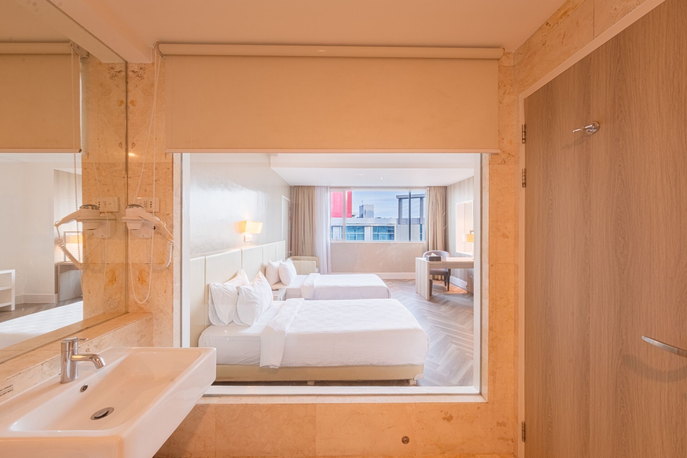Quarto de hotel com um espelho grande, pia, cama com roupa de cama branca e vista para a cidade pela janela.